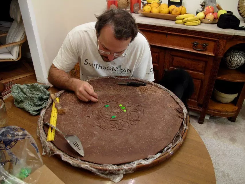 Eric Hollinger sitzt an einem kleinen Holztisch, der sich über eine große runde Platte fester Milchschokolade beugt.