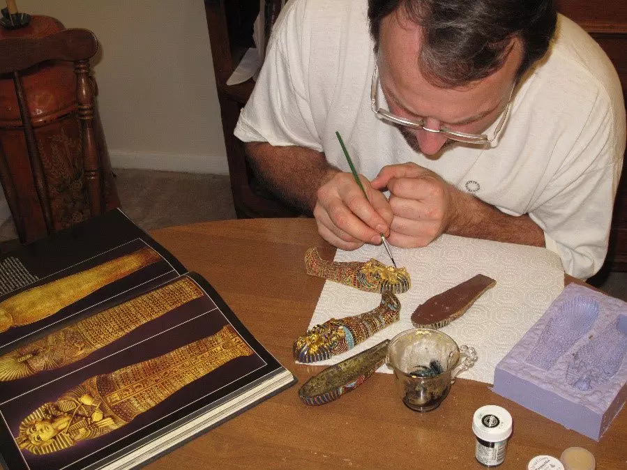 Ерик Холингер седи за малим дрвеним столом и слика јестиво злато на малом чоколадном саркофагу.