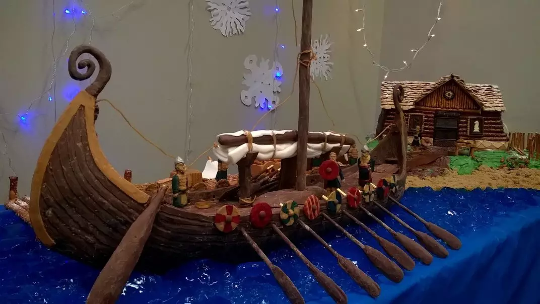 Smeđa, čokoladna rekreacija vikinškog broda zajedno sa veslima usidrenim blizu smeđe, čokoladna torta rekreacija vikinškog doma izložena na pravokutnom stolu s plavim stolnjakom.
