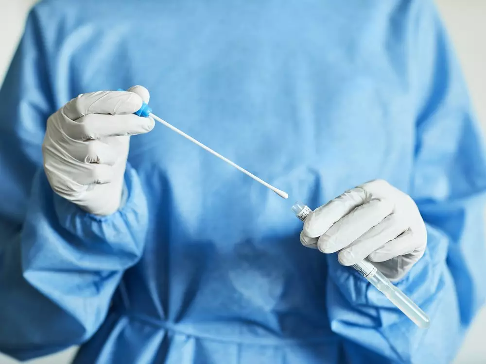 Một nữ bác sĩ mặc áo choàng phẫu thuật màu xanh lam cầm một chiếc tăm bông dài dùng để xét nghiệm covid-19