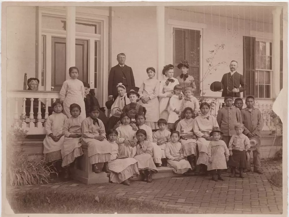 Sepijos atspalvio atvaizdas jaunų moterų su keletu jaunų berniukų ir merginų, sėdinčių priešais baltą namą verandoje, vaizdas. Pratas stovi tamsiais drabužiais už grupės