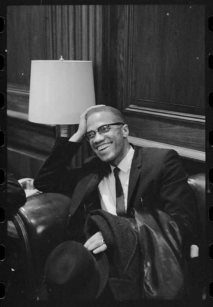 Malcolm X, yon nonm wo mens ak linèt, mete yon kostim ak yon kravat epi li mete tèt li apiye sou bra l pandan l ap ri.