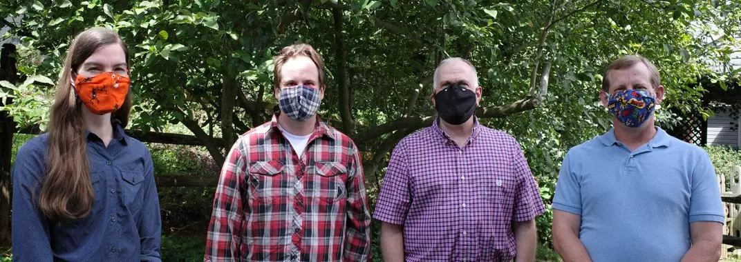 Štirje ljudje, vsi z maskami, stojijo pred zelenimi drevesi in se nasmehnejo nekaj metrov drug od drugega