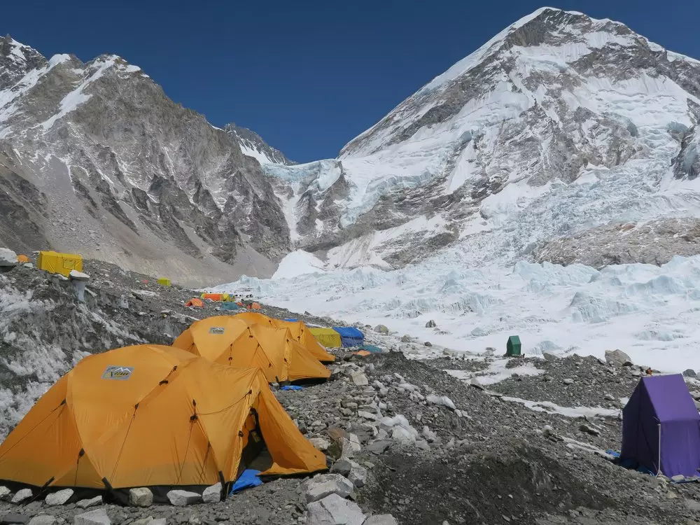 Ett foto av Everest Base Camp. Tält i olika färger ligger utspridda över den grå, steniga marken. Snötäckta berg är i bakgrunden.