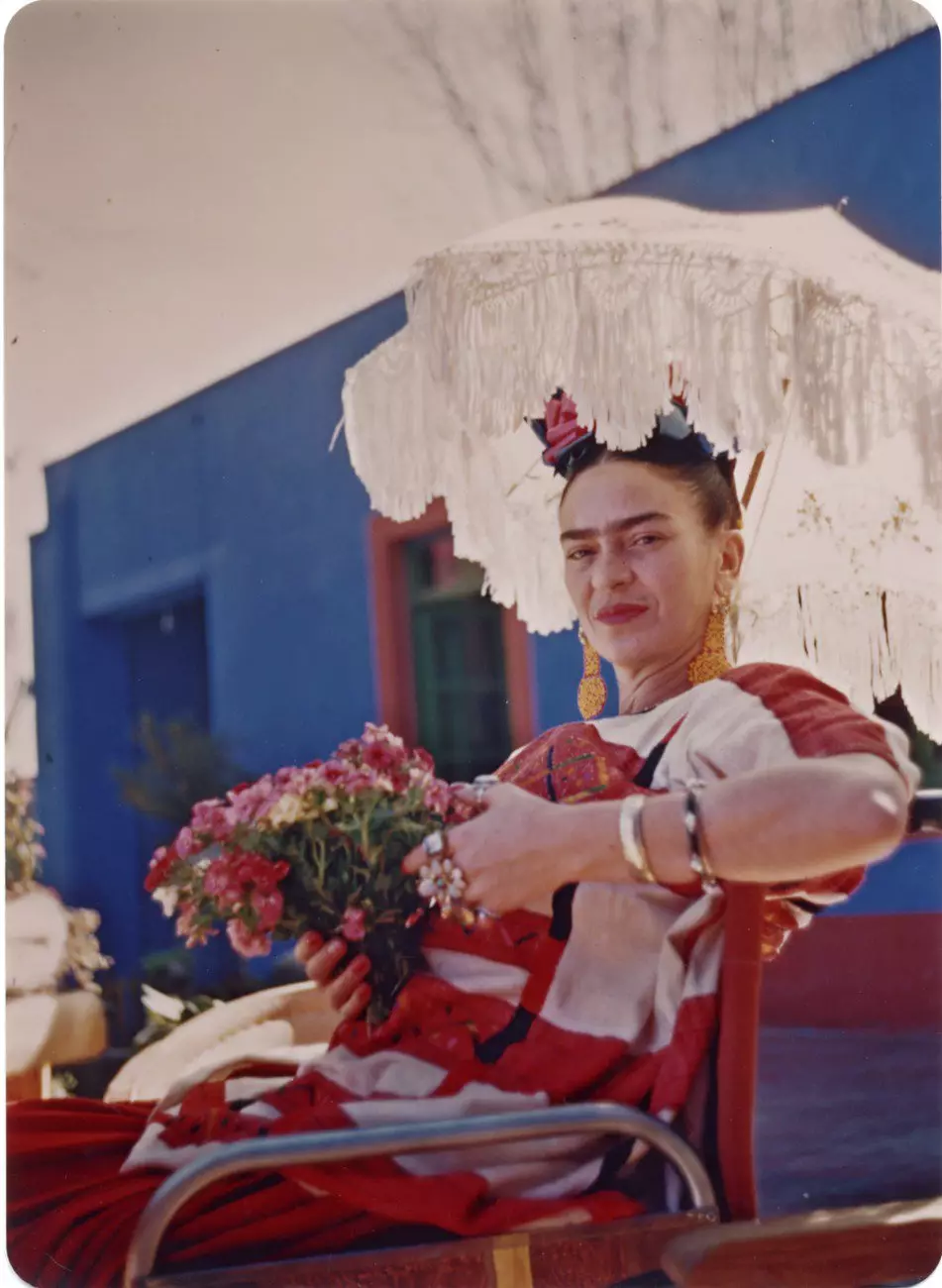 Kahlo 拿着一束鲜花，戴着花冠和红白相间的连衣裙，坐在她明亮的蓝色房子前的白色阳伞下