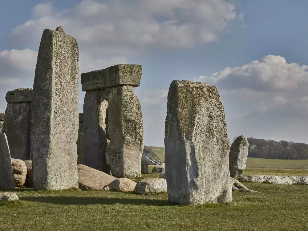 Una vista de les emblemàtiques pedres quadrades de Stonehenge, sota un cel blau amb núvols blancs inflats; a la distància entre dues de les grans pedres, una carretera transitada plena de cotxes serpenteja sobre un turó