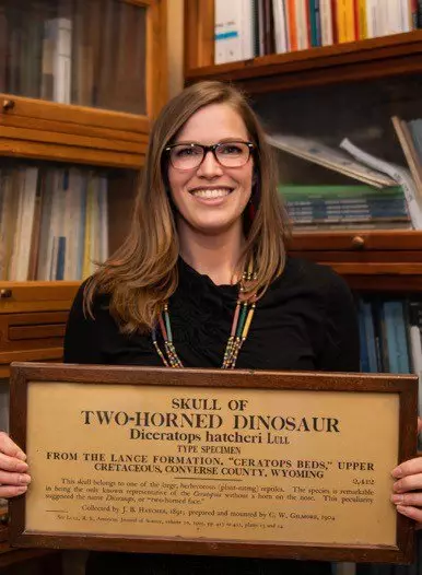Diana Marsh nyepeng labél paméran kai ti aula fosil awal Smithsonian kalayan rak buku di tukangeunana.