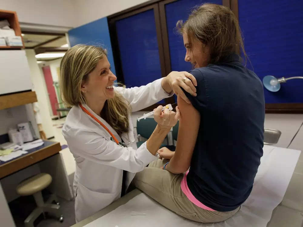 Hình ảnh một bác sĩ nhi khoa đang tiêm vắc xin HPV cho một bé gái 13 tuổi trong phòng khám của bác sĩ. Cả hai đối tượng đang mỉm cười với nhau.
