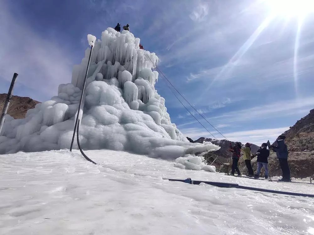 Ladakh Valley의 밝은 날, 한 팀의 사람들이 얼음 사리탑에서 작업합니다. 그것은 눈 덮인 땅 위에 앉아 있는 키가 큰 원뿔 모양의 얼음 더미입니다.
