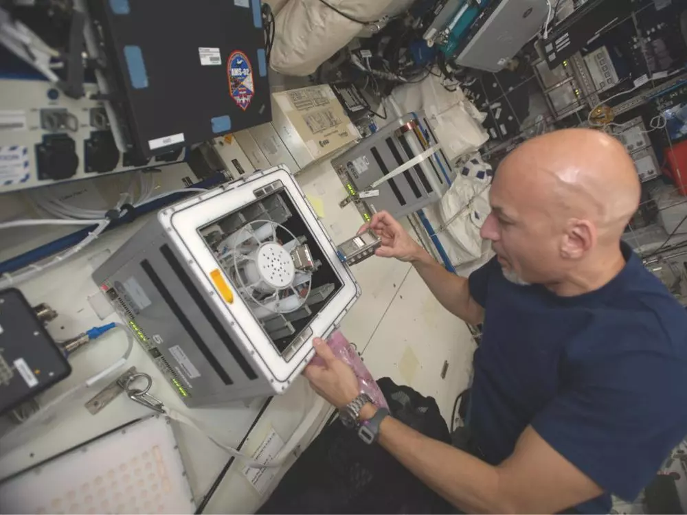 Phi hành gia mặc áo phông xanh dương đặt một lọ nhỏ vi khuẩn và đá bazan vào máy ly tâm trên trạm vũ trụ quốc tế