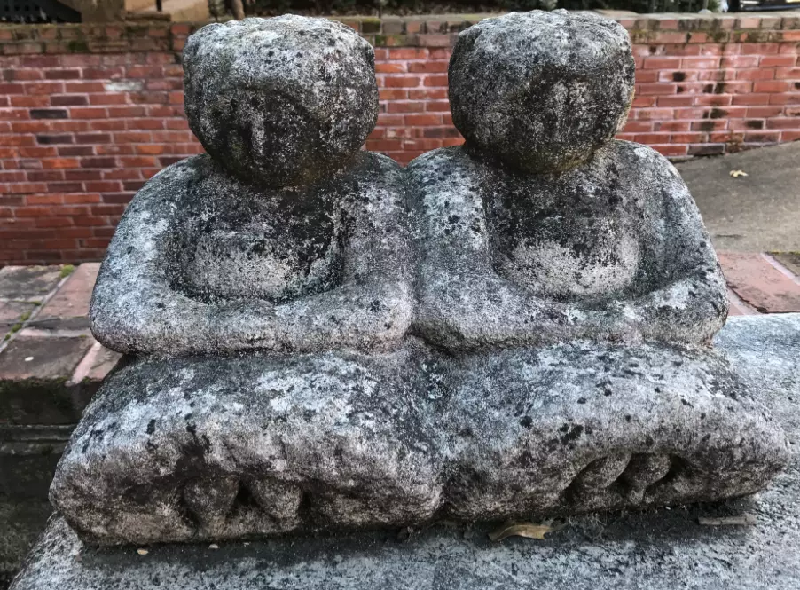 Sebuah patung dua wanita duduk, tertutup lumut, tanah dan lapuk oleh waktu, terletak di luar di depan trotoar dan dinding bata