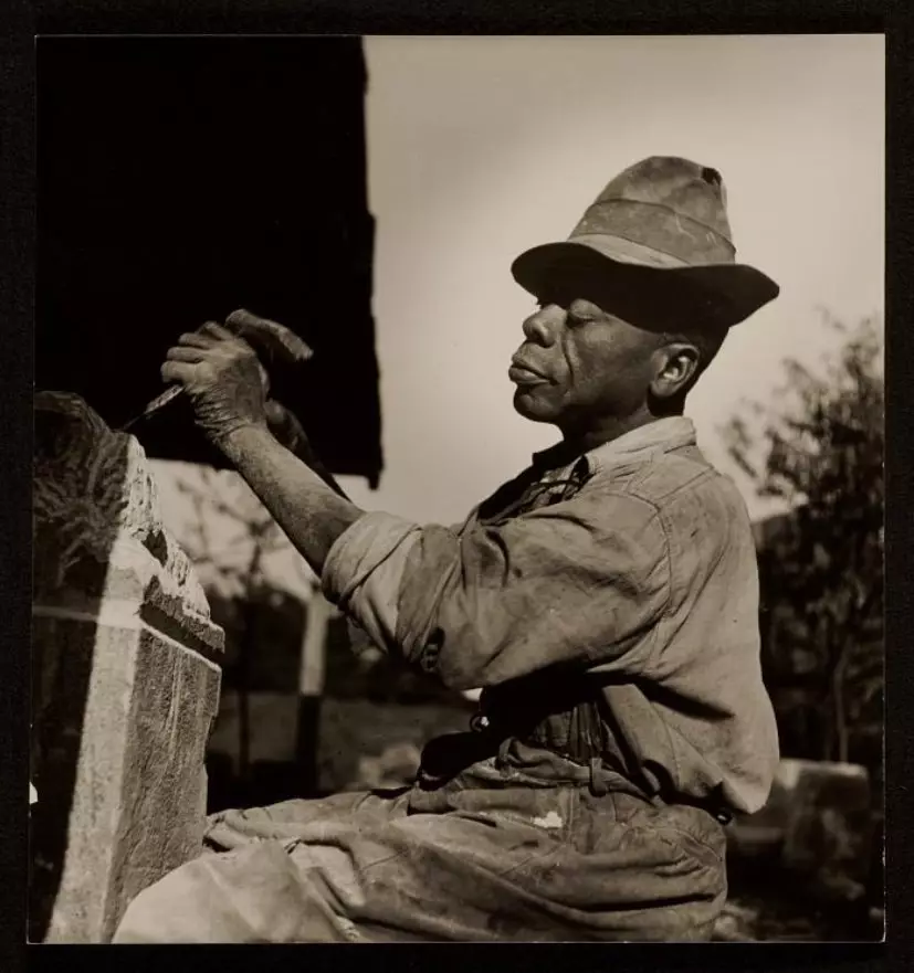 En svart man i hatt, overall och skjorta sätter sig och arbetar intensivt på en skulptur