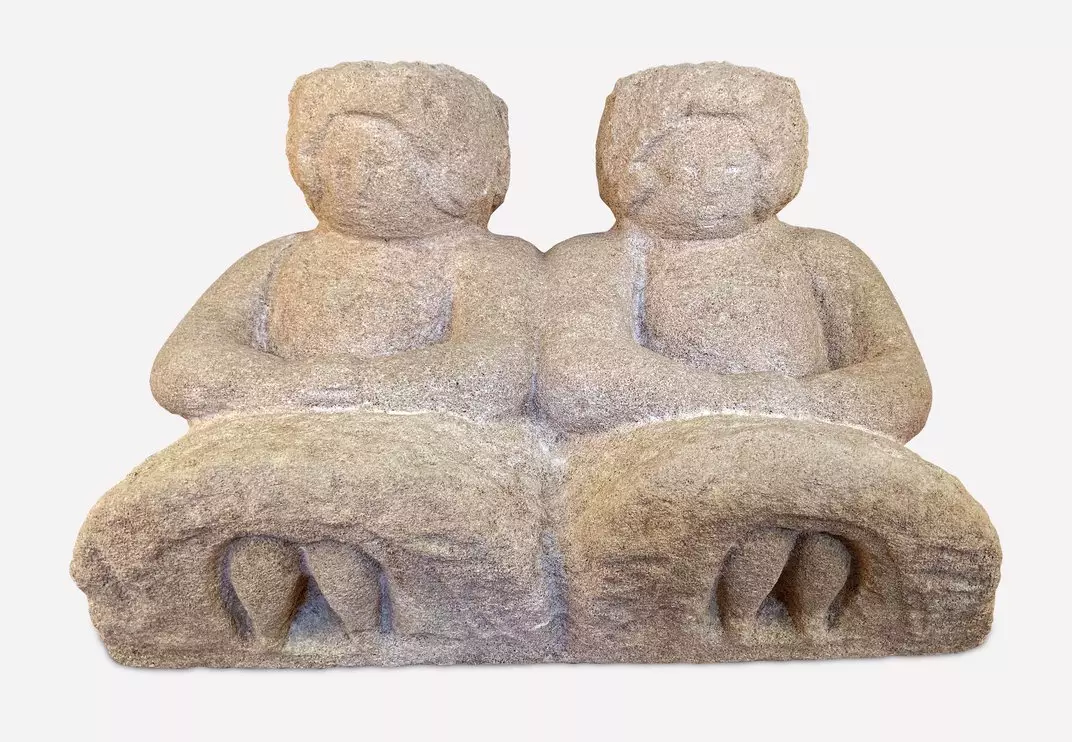 Une vue d'une sculpture en pierre blanchâtre de deux femmes, assises côte à côte avec leurs petits pieds sortant de leurs robes identiques
