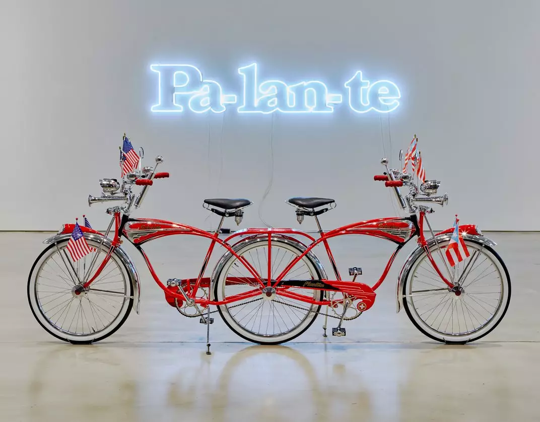 Một tác phẩm điêu khắc của một chiếc xe đạp màu đỏ. Nó có hai đầu phía trước, nối với nhau ở phía sau. Phía trên nó, một bóng đèn neon cho biết