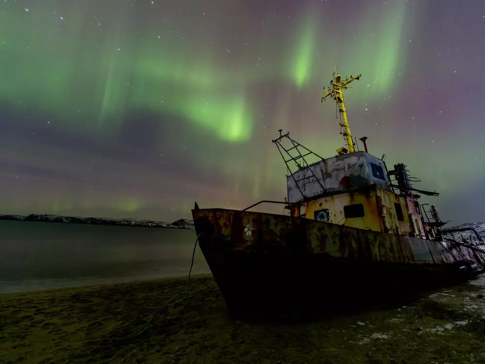 Slika severnega sija na nebu. Fotografija prikazuje tudi dotrajano ladjo, ki sedi na kopnem ob vodi.