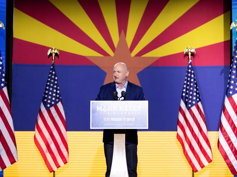 Mark Kelly berdiri di atas panggung di podium di depan bendera Arizona