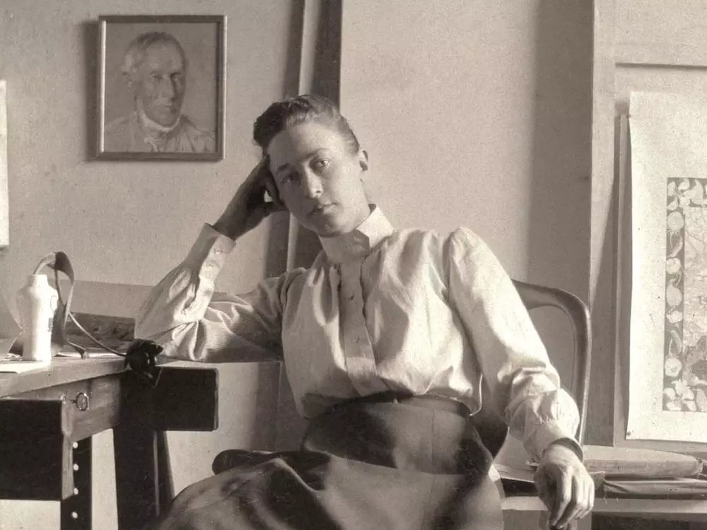 הילמה אפ קלינט, אישה לבנה בחולצה לבנה וחצאית שחורה ארוכה, משעין את ידה על מרפק אחד ויושבת ליד השולחן בסטודיו שלה.