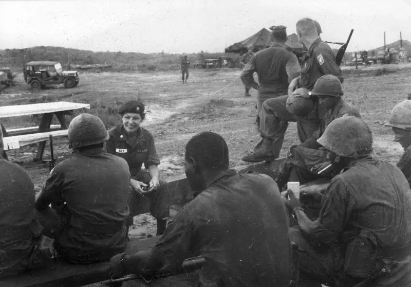 Martha Raye i samtale med medlemmer af den 173. luftbårne brigade. Raye og soldaterne sidder udenfor på bænke. Telte, en jeep og andre soldater kan ses i det fjerne i baggrunden.