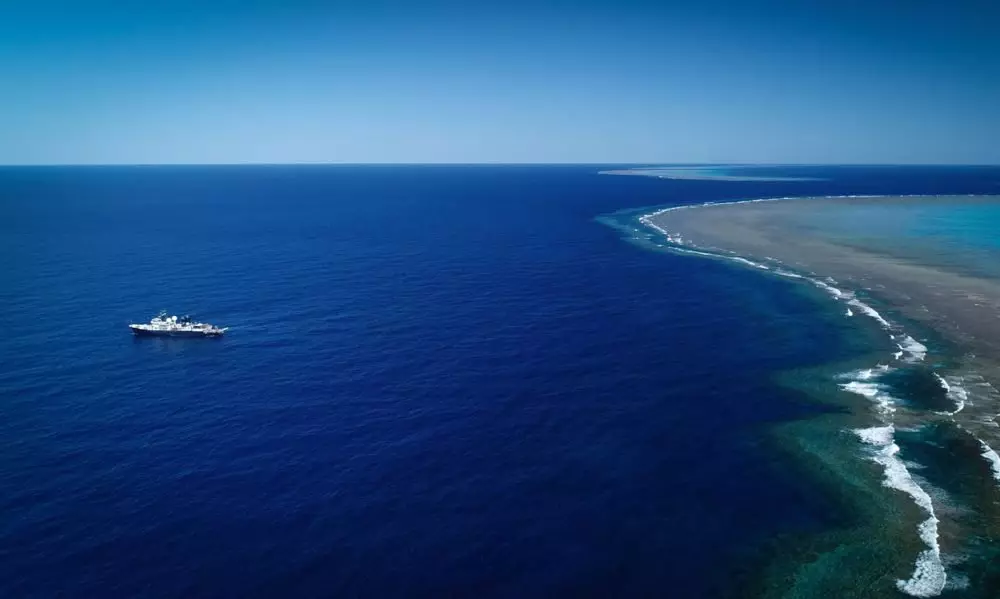 Фотография, сделанная с высоты (вероятно, с помощью дрона), показывает огромный ярко-синий океан. Исследовательское судно находится слева. Длинный зелено-бежевый участок мелководных коралловых рифов находится с правой стороны. Горизонт, где глубокий синий океан встречается с светом