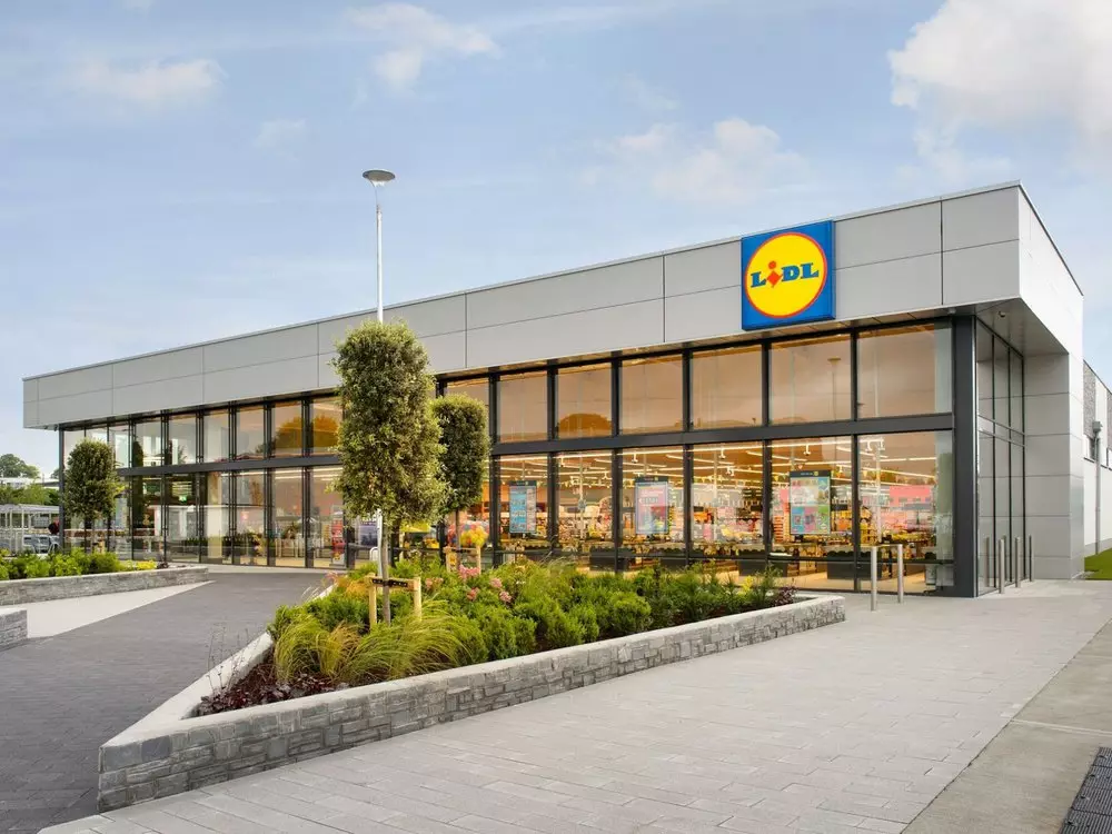 An image of a brightly lit new Lidl store; a large square building with the store's logo, a blue square with a yellow circle and blue and red block text that reads LIDL; surrounded by pavement and a parking lot
