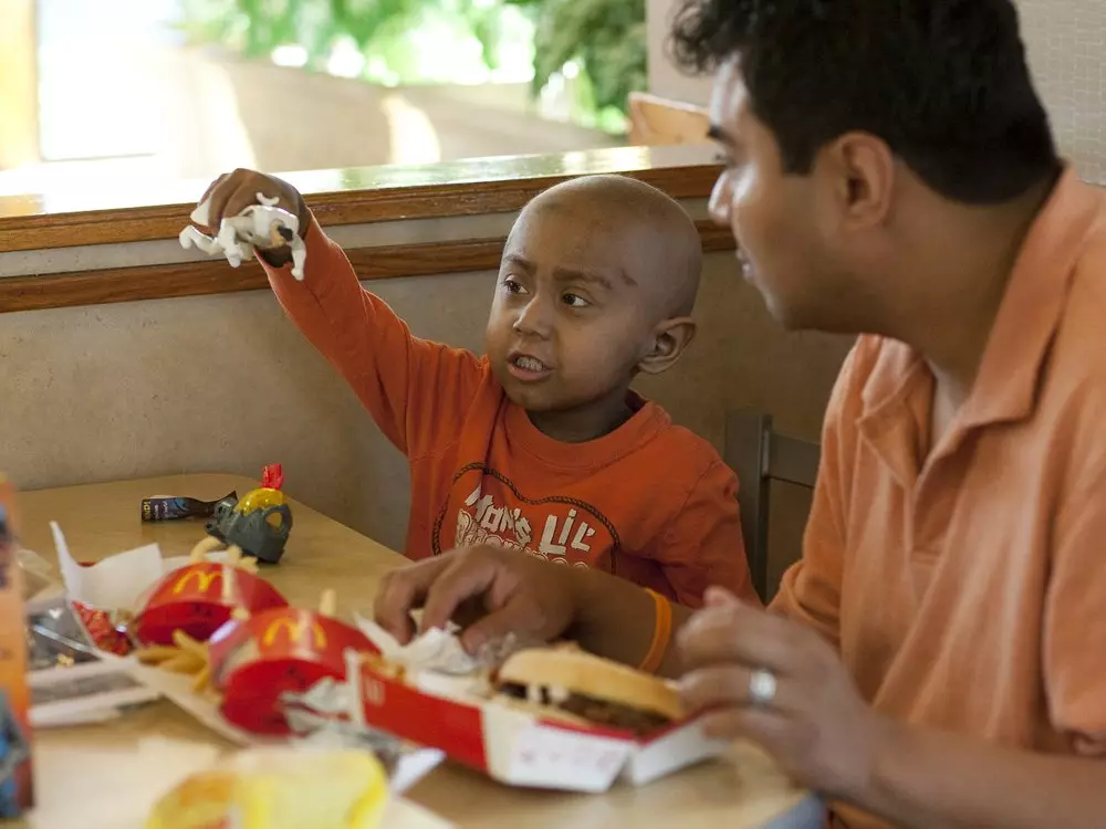 Egy 5 éves gyermek képe, aki egy McDonald's Happy Meal játékkal játszik. A gyerek előtt egy boldog étkezés sült krumplival és a gyerek mellett ül az apja.