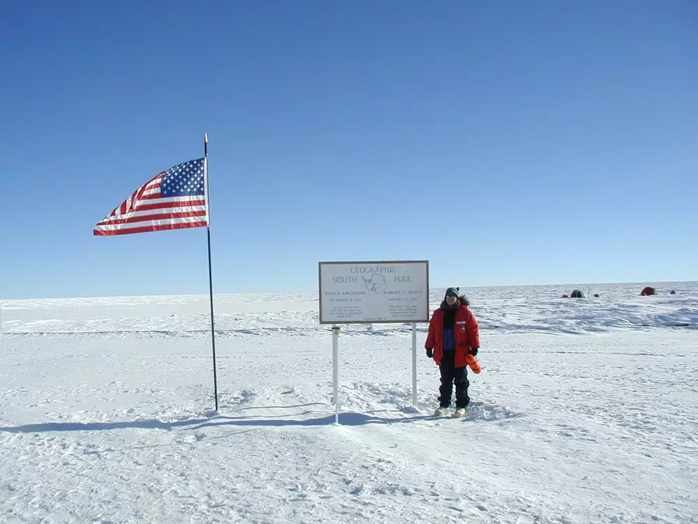 ژئولگیست کری کوریگان برای عکس گرفتن در برف در کنار پرچم آمریکا و تابلویی که روی آن نوشته شده است.