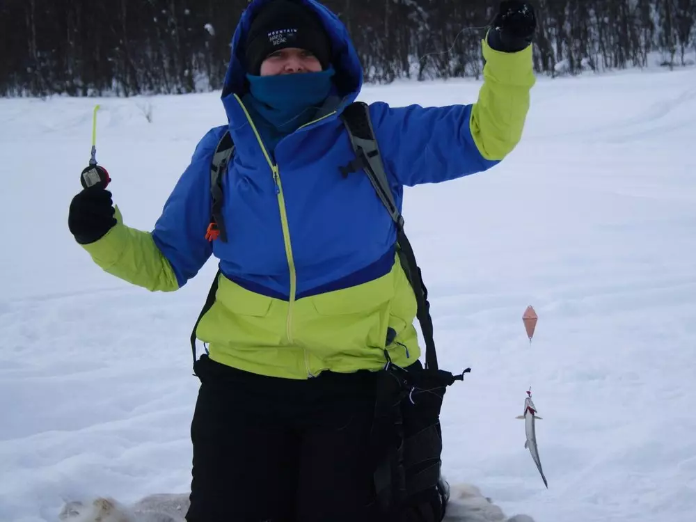 Persona, kas valkā ziemas rīkus, nometusies ceļos sniegā, turot augšā makšķerēšanas auklu ar mazu zivtiņu