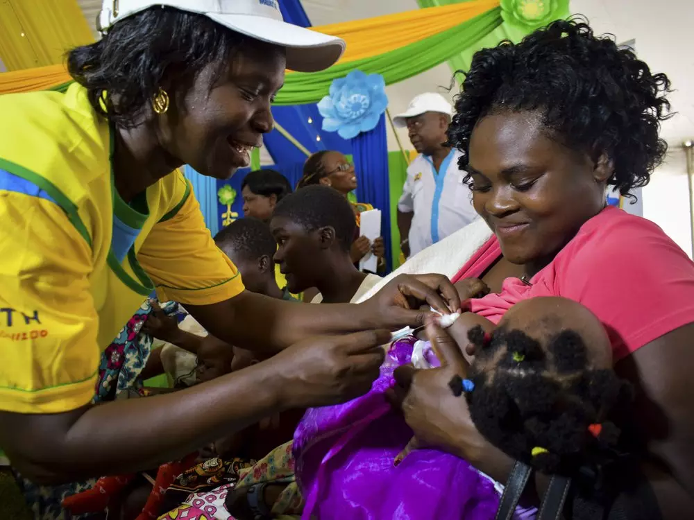 કેન્યામાં મેલેરિયા સામે બાળકને રસી આપતા આરોગ્ય કાર્યકર. બાળકને તેની માતાએ પકડી રાખ્યું છે જે હસતી છે.