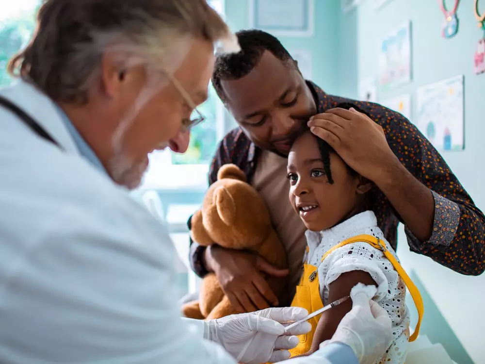 Слика на постар машки лекар кој вакцинира мало дете. Детето е насмеано и го држи нејзиниот татко.