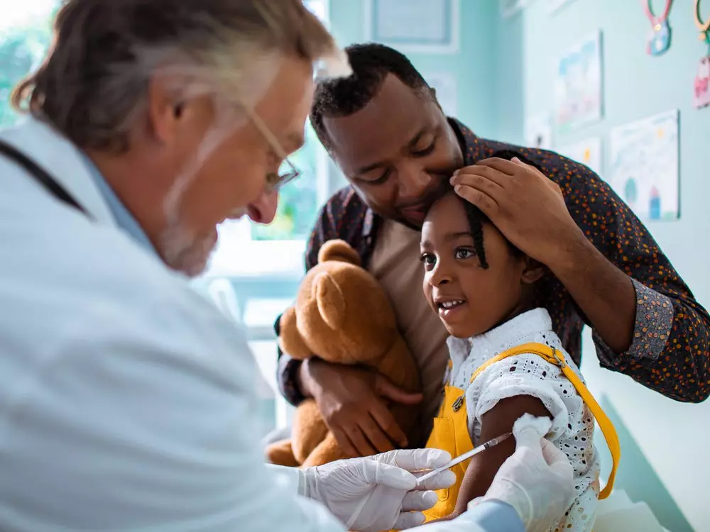 In byld fan in âldere manlike dokter dy't in jong bern faksinearret. It bern glimket en wurdt holden troch har heit.