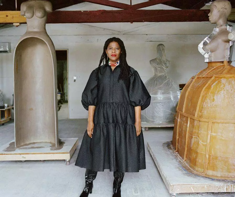 Simone Leigh, unha muller afroamericana, leva un vestido negro voluminoso e está diante de tres das súas esculturas, que son representacións abstraídas de figuras femininas encima de saias de aro e xerras; Leigh enfróntase á cámara cunha expresión seria