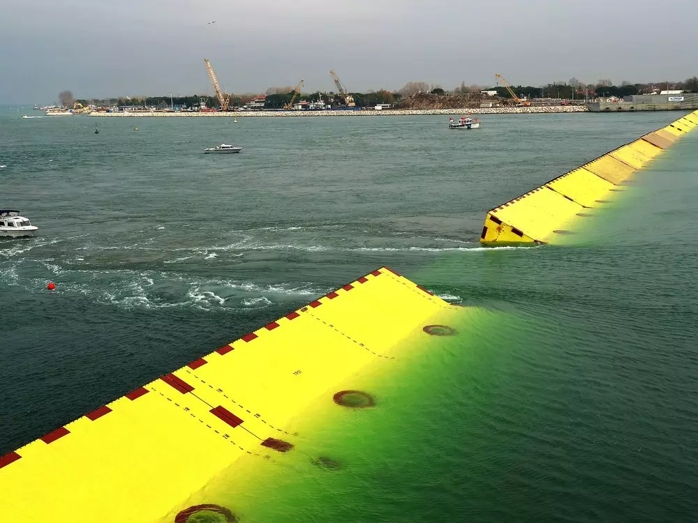 Vicino alla riva, le barriere antiallagamento emergono dall'acqua. Sono grandi, di colore giallo brillante e di forma triangolare per deviare l'acqua. Ci sono diverse barche in acqua nelle vicinanze. Sullo sfondo c'è il Lido, un'isola barriera vicino a Venezia.