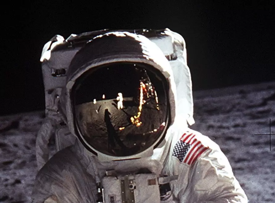 Astronaut Buzz Aldrin, keď stojí na Mesiaci, nosí veľkú prilbu a biely vesmírny oblek. V jeho prilbe sa odráža americká vlajka, Neil Armstrong a ďalší mesiac. Kamenný sivý povrch Mesiaca tvorí pozadie.