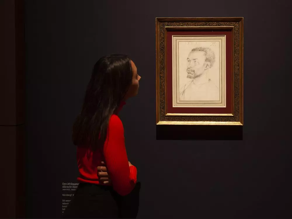 A person with long dark hair stands in a darkened gallery and looks at an illuminated white paper sheet, with a sketch of an African man's head and shoulders