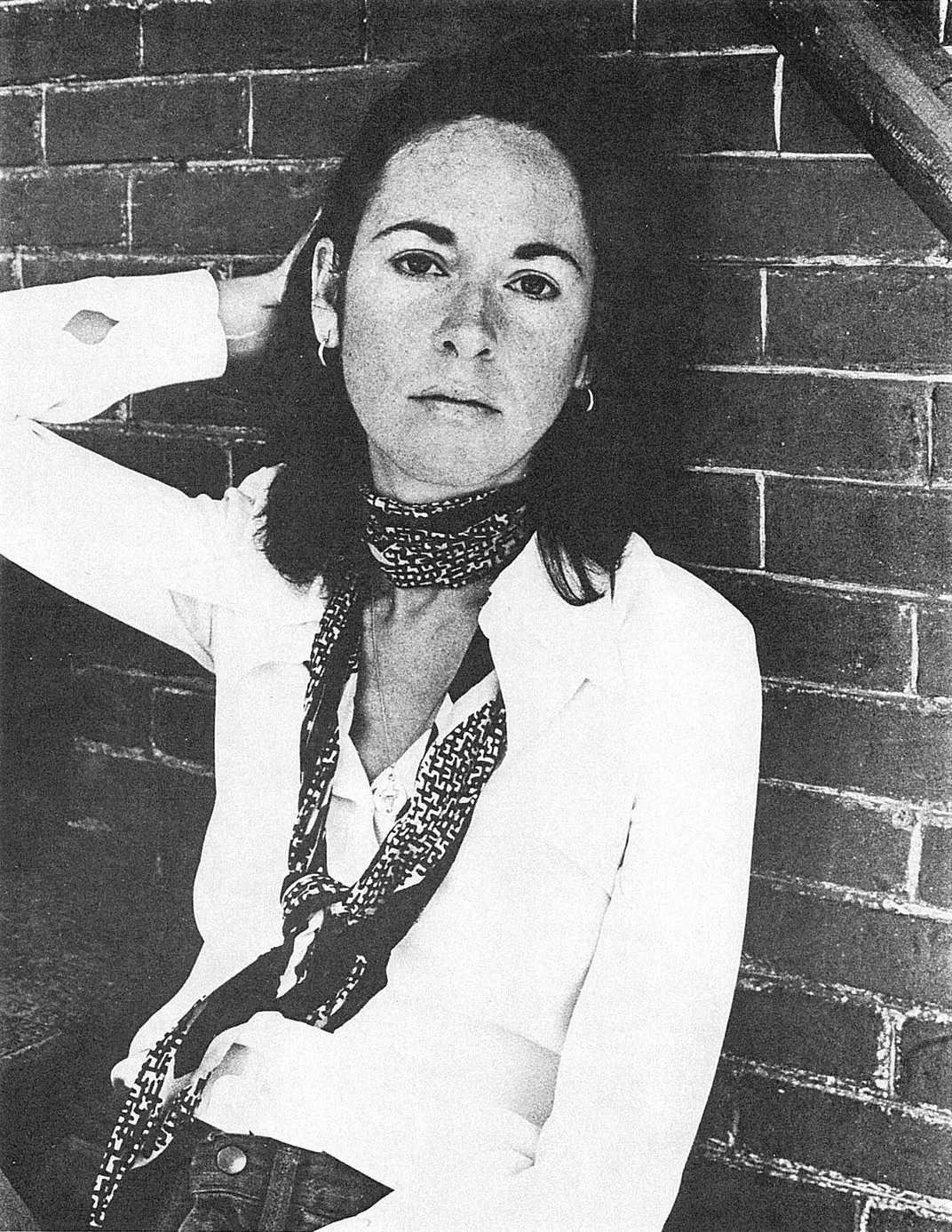 Een jonge, elegante Gluck leunt tegen een bakstenen muur in een zwart-wit afbeelding; houdt een hand achter haar hoofd in een pose, kijkt serieus naar de camera; in een wit overhemd met schouderlang donker haar, oorbellen en een dunne sjaal losjes om haar nek gebonden