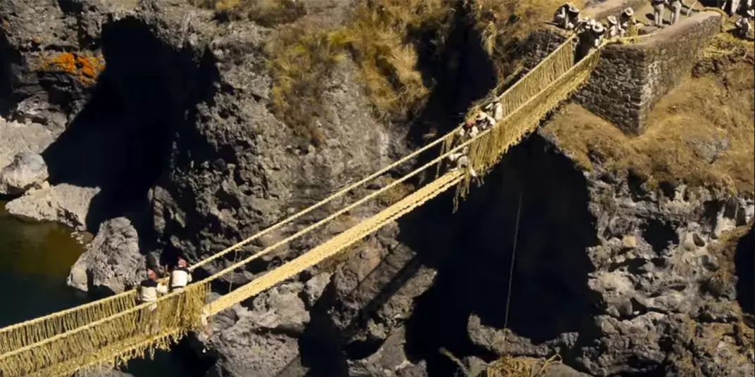Fotografiat des de dalt, el pont inacabat de la riba del riu. Els cables de terra i els cables manuals estan al seu lloc. Dos grups d'homes treballen des de cada extrem del pont teixint els costats.