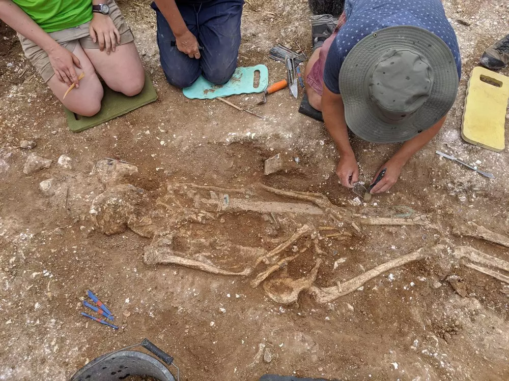 Trys žmonės atsiklaupia ir pasilenkia pažiūrėti į kapą; atrodo, kad vienas skrybėlę nešiojantis asmuo švelniai liečia arba pašalina dalį skeleto