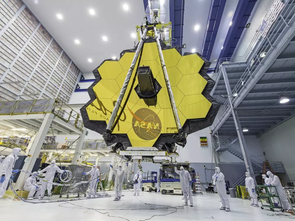 Imej Teleskop Web James di makmal NASA. Kelihatan juruteknik berdiri di bawahnya manakala cermin telekop berbentuk madu tergantung di atas. Logo NASA dilihat terpantul di cermin.