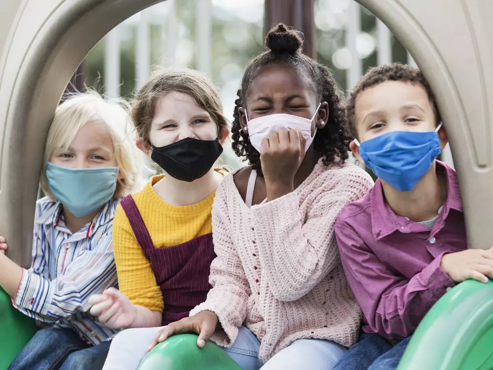 Een diverse groep gemaskerde kinderen zit bovenaan een glijbaan in een park