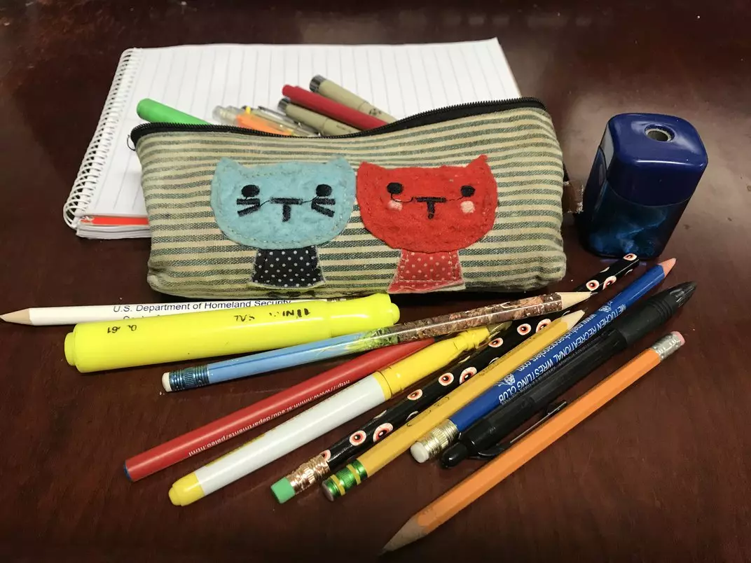 एक लकड़ी की मेज पर कई पेंसिल, पेन और हाइलाइटर के साथ एक नोटबुक, पेंसिल होल्डर और पेंसिल शार्पनर।