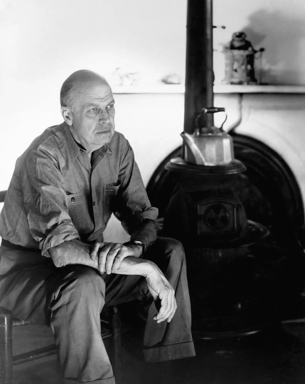 Et sort/hvid billede; ældre Hopper sidder, for det meste skaldet, og drejer lidt fra kameraet til venstre foran et gammeldags komfur og elkedel; han tager fat i den ene arm med den anden hånd og bærer en enkel skjorte og bukser med knapper; bærer et seriøst udtryk