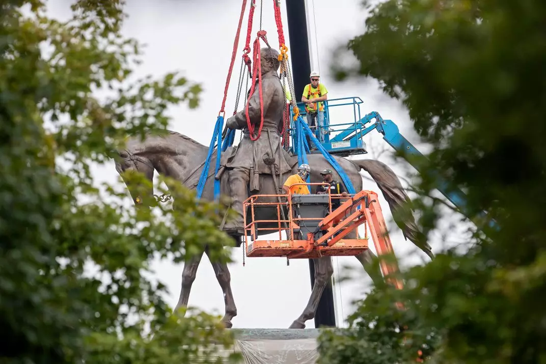 Entre as árvores, uma vista de um guindaste de construção laranja brilhante guiando a grande estátua para o chão