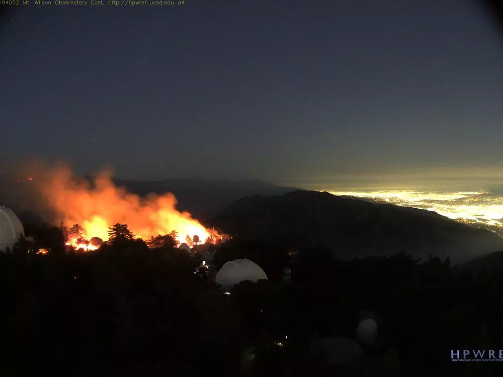 Панорамна снимка на мекото сияние на Пасадена и Лос Анджелис вдясно, шир от тъмни нощни хълмове, два бели куполни телескопа вляво с високи, ярко оранжеви и жълти пламъци, избухващи наблизо, драматични на фона на мрака