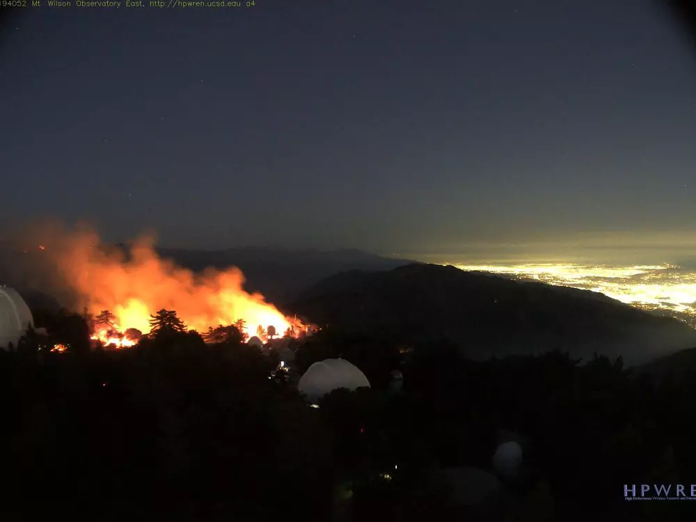 Isithombe se-panoramic sokukhanya okuthambile kwe-Pasadena ne-Los Angeles kwesokudla, umkhathi wamagquma amnyama ebusuku, izibonakude ezimbili ezibuswe ngokumhlophe ngakwesobunxele ezinamalangabi amade, awolintshi akhanyayo naphuzi aqhuma eduze, amangalisayo ngokumelene nobumnyama.