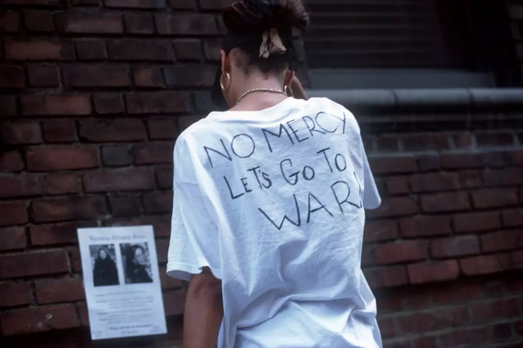 Ženska je obrnjena stran od kamere in gleda plakat pogrešane osebe 13. septembra. Nosi belo srajco z ročno napisanim sporočilom NO MILOSTI, GREMO V VOJNO