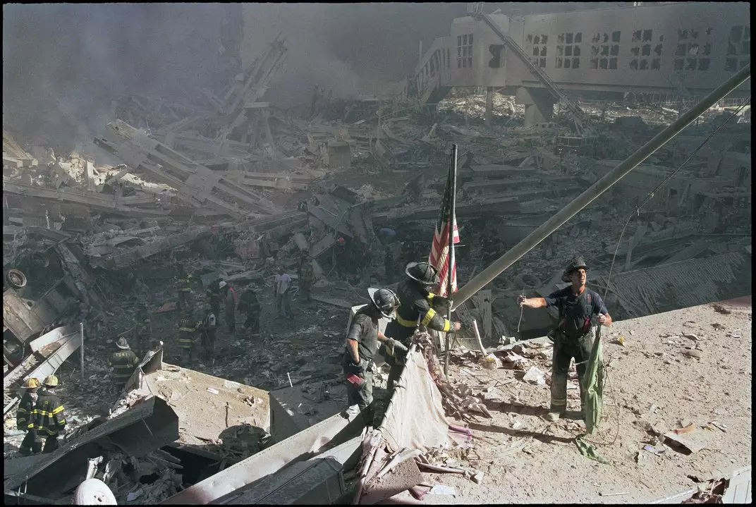 Sekelompok tiga pria berdiri dalam cahaya kuning senja, menebarkan bayangan di belakang mereka, mengibarkan bendera di depan reruntuhan yang terhampar luas.