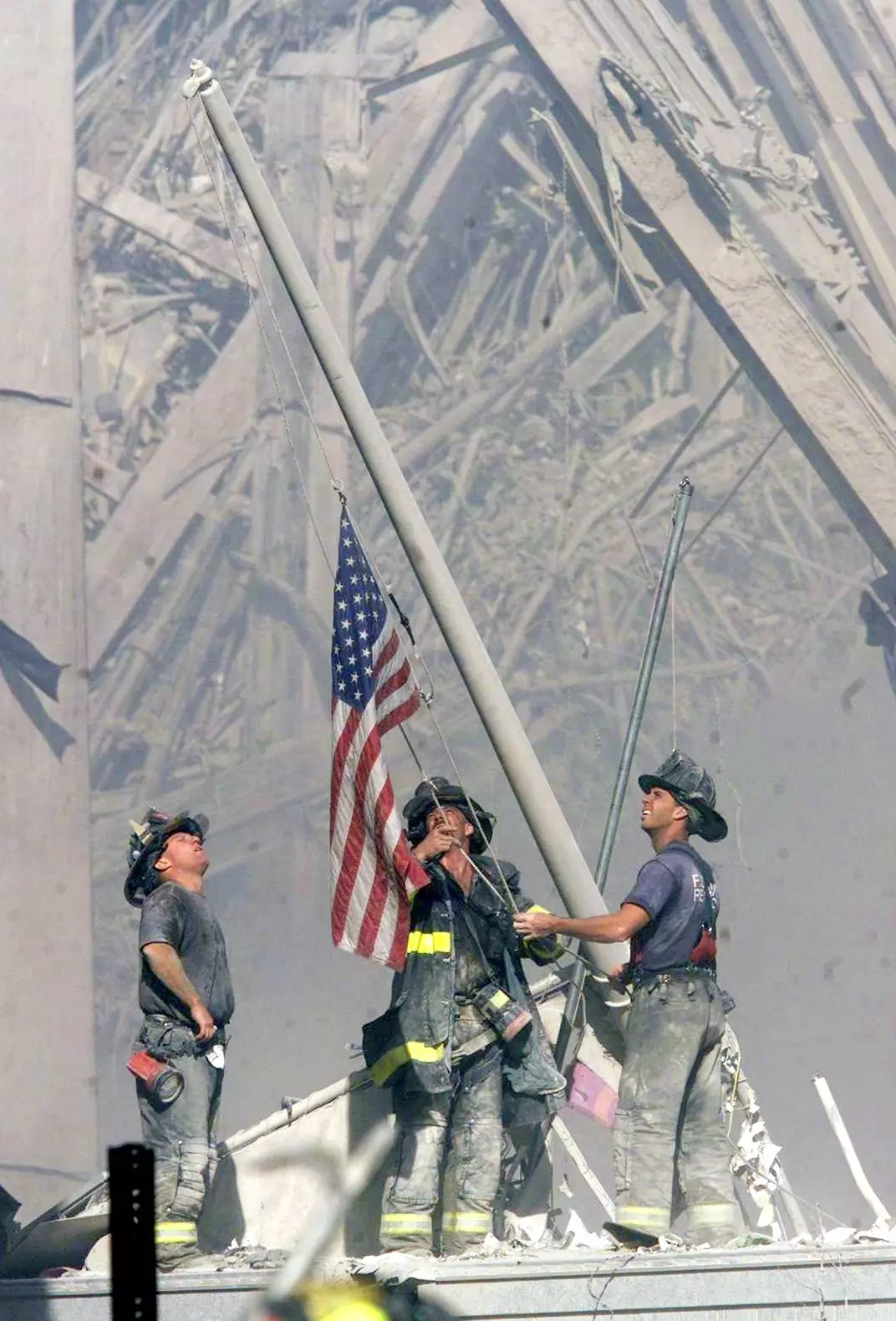 'n Vertikale skoot van drie wit mans wat besig is om 'n Amerikaanse vlag op 'n getitelde vlagpaal te hys, wat uit die puin van Ground Zero steek