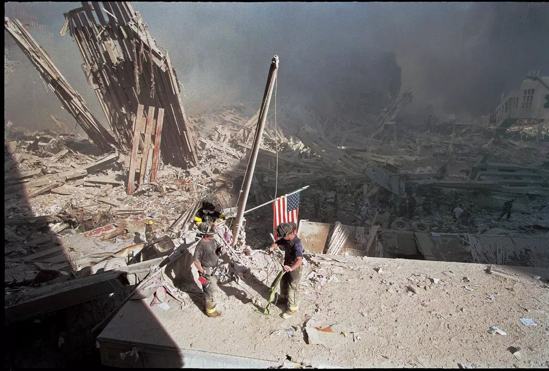 Vaakasuora laaja näkymä kohtauksesta, jossa on hylkyjä ja savua niin pitkälle kuin silmä näkee; keskellä kolme pientä palomiestä poistavat Yhdysvaltain lipun tangosta ja sitovat sen suureen lipputankoon