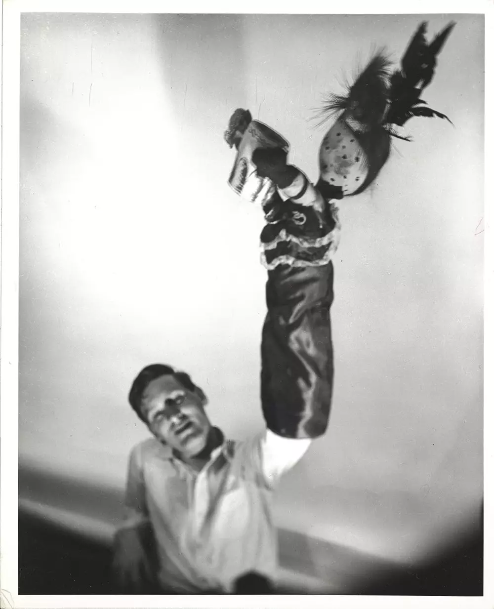 Photo floue en noir et blanc d'un homme assis sur une chaise portant une chemise blanche et tenant une grande marionnette qui ressemble à un oiseau.