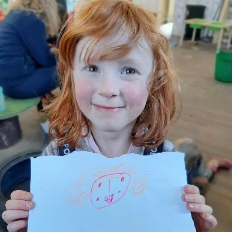 Unha rapaza pálida e pelirroja con flequillo sorrí e sostén un anaco de papel diante de si, co que parece un debuxo do rostro sorrinte dunha persoa e o pelo rizado laranxa.